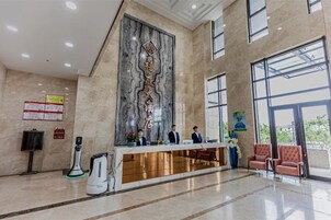Área de recepção de hotel com parede de mármore, balcão de atendimento, espaço para sentar e grandes janelas.