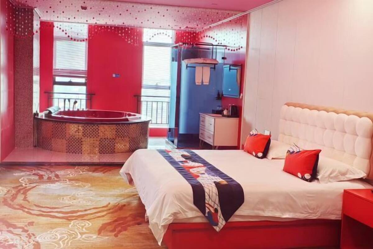 Quarto com cama, mesa de cabeceira vermelha, cabeceira branca, tapete com padrão vermelho e branco, banheiro pequeno e varanda com corrimão vermelho.