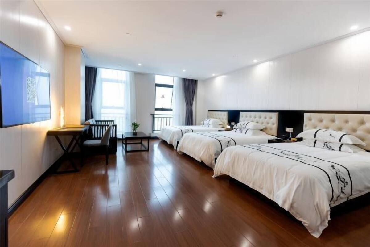 Quarto de hotel com piso de madeira, duas camas com roupa de cama branca, uma escrivaninha com cadeira e uma TV fixada na parede.