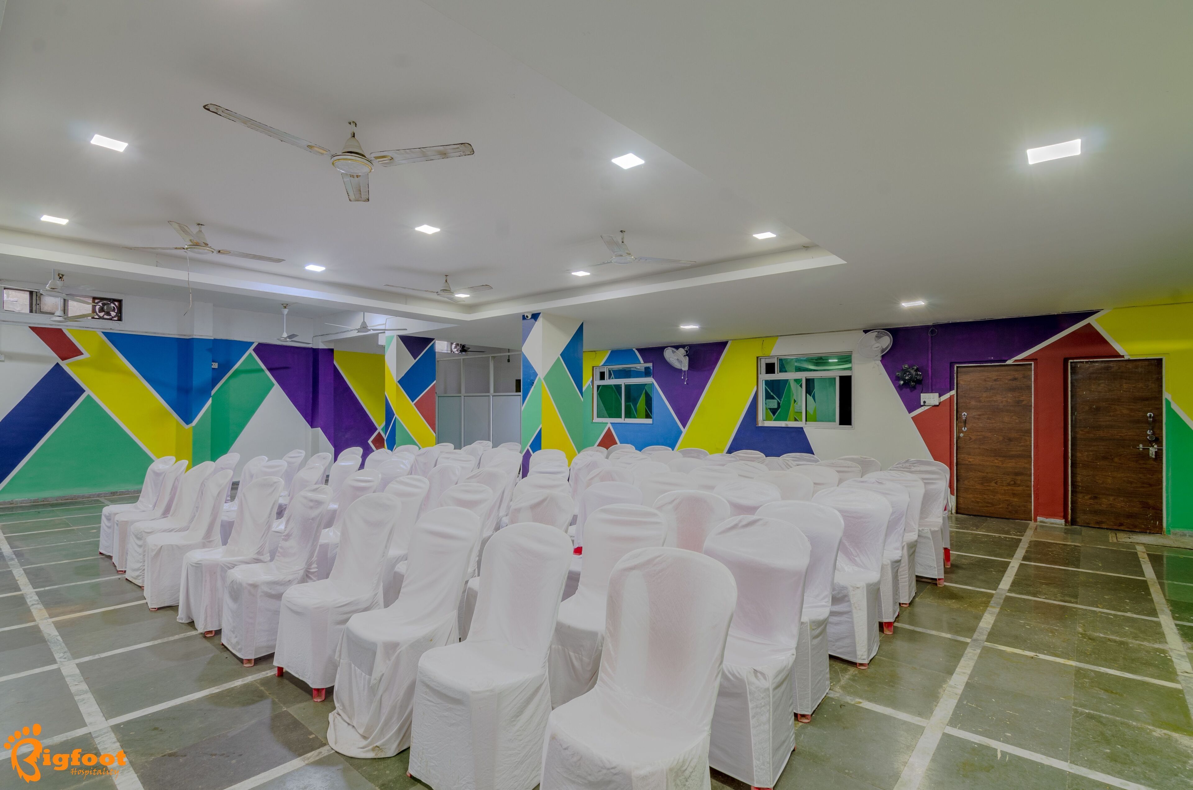 Uma sala de conferência com fileiras de cadeiras cobertas de branco, ventiladores de teto e designs geométricos coloridos nas paredes.