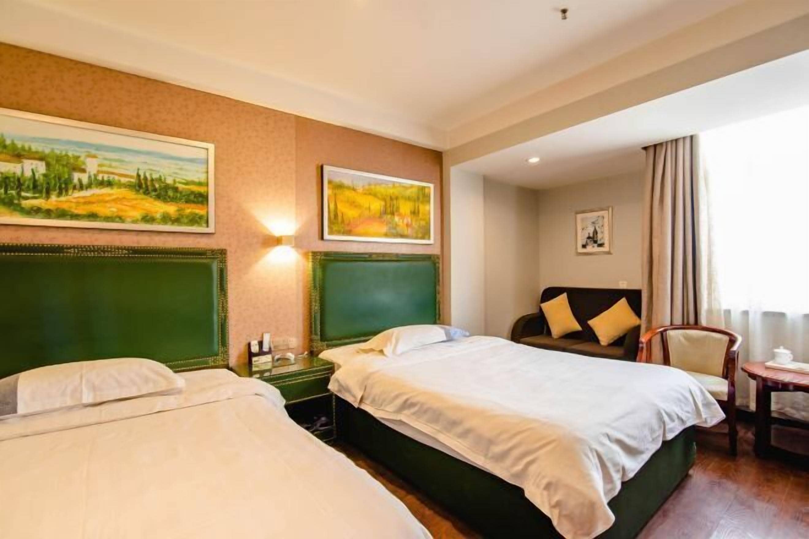 Quarto de hotel com duas camas, cabeceira verde, mesa de cabeceira, uma cadeira, uma mesa e duas pinturas na parede.