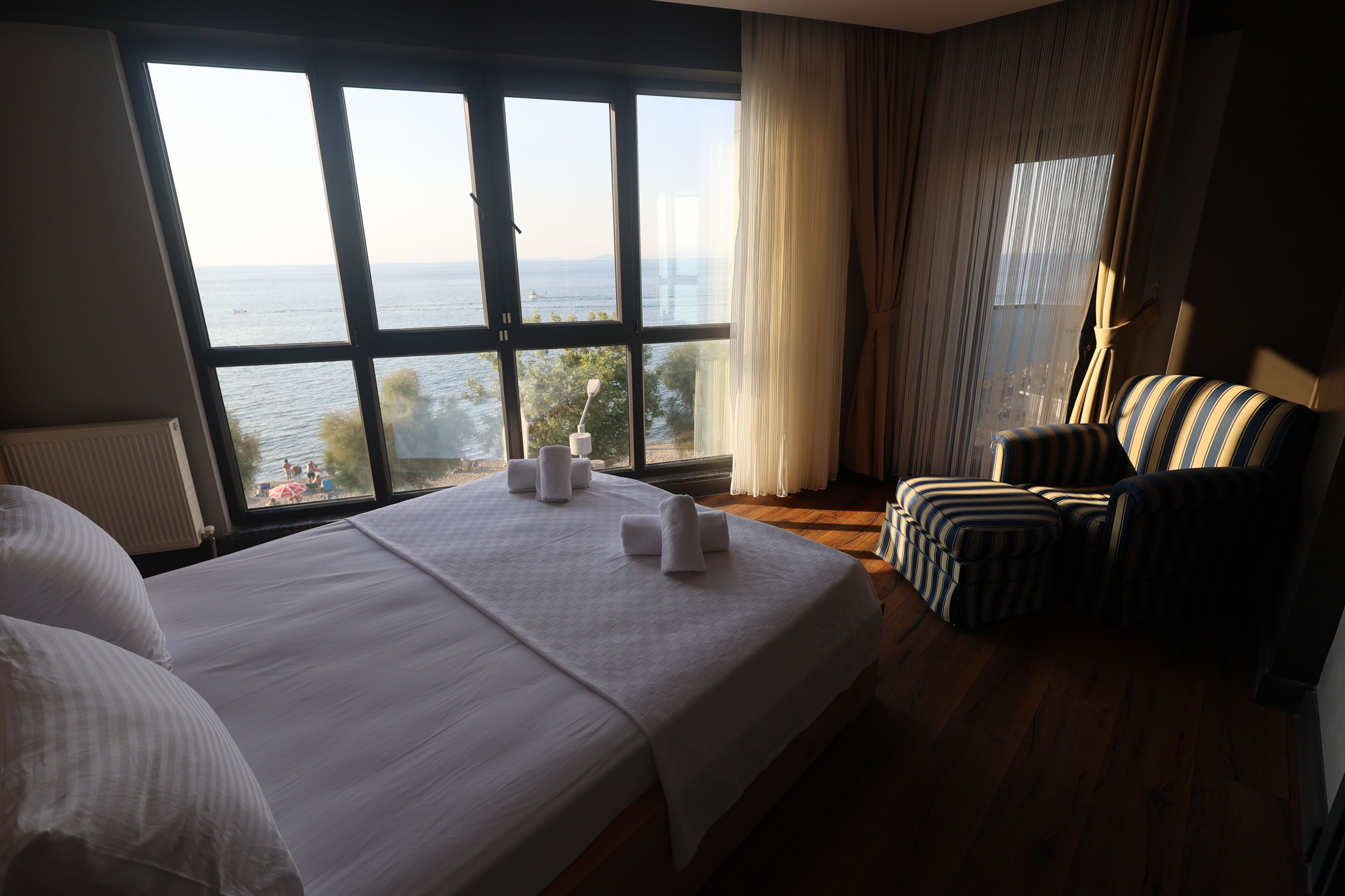 Quarto de hotel com uma grande janela, uma cama com roupa de cama branca, uma poltrona listrada e vista para o mar.