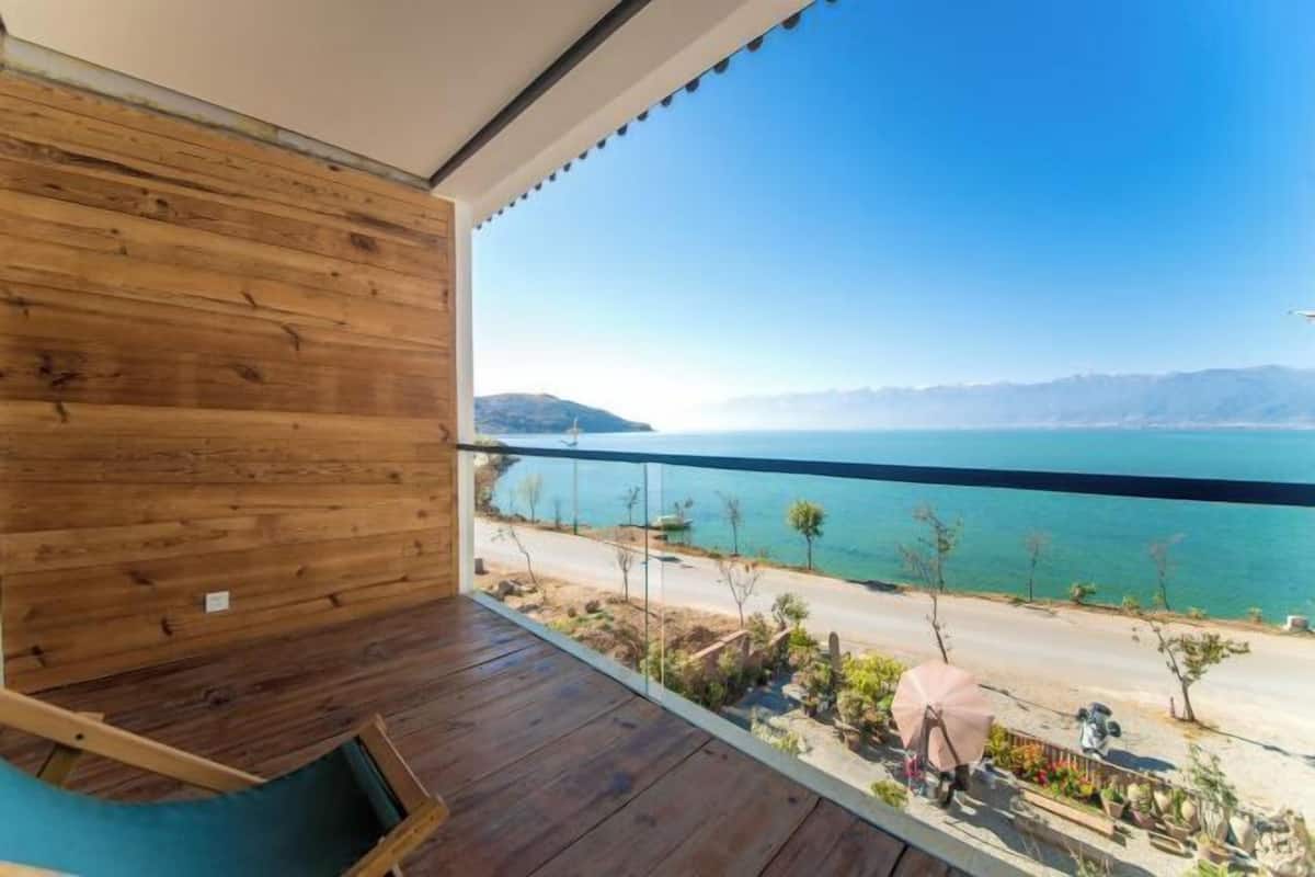 Uma varanda com piso de madeira, uma cadeira e vista para o mar e montanhas.