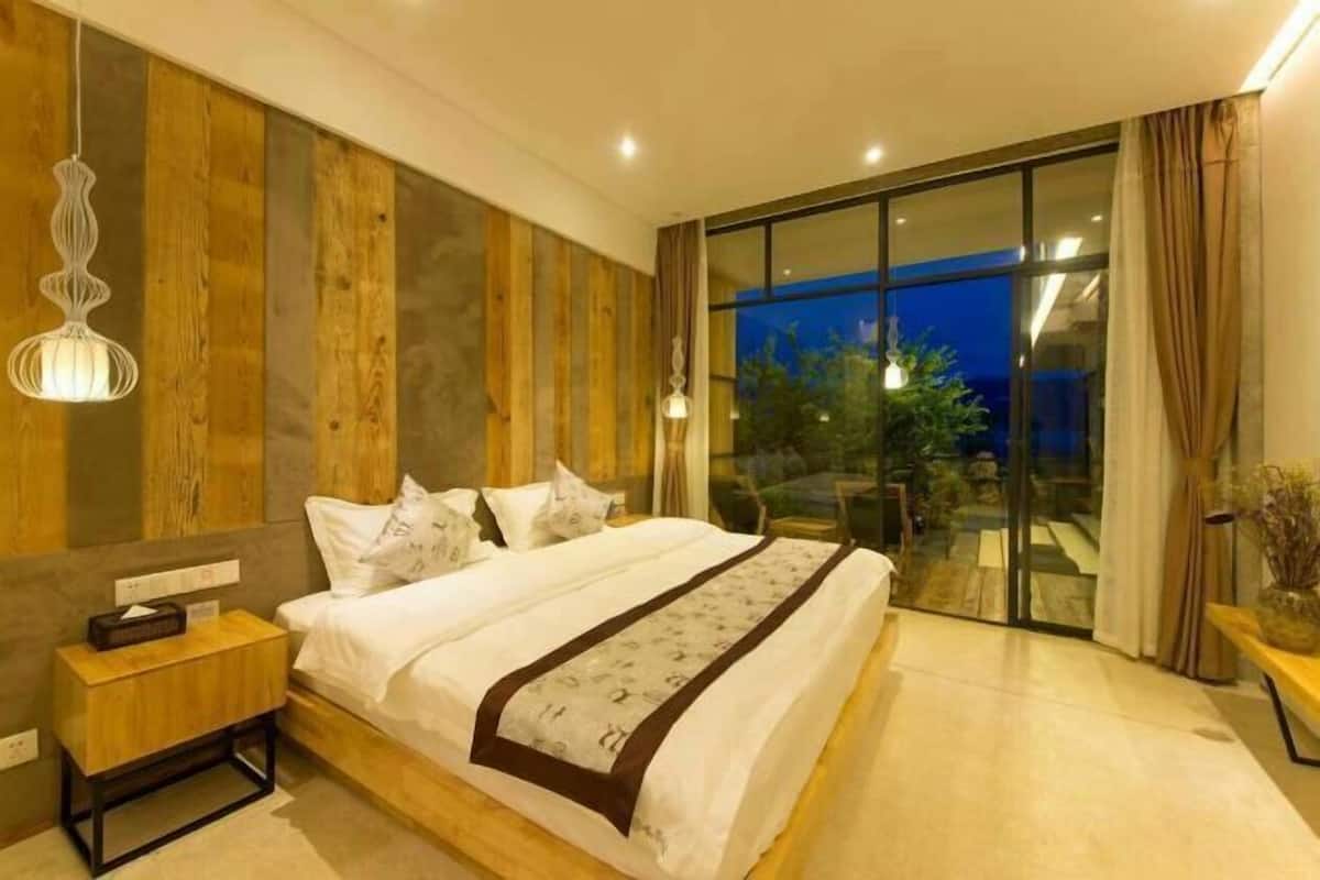 Quarto moderno com uma cama grande, painéis de madeira e vista do exterior através de uma porta de vidro.