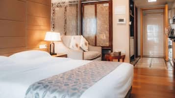 Quarto de hotel com uma cama, mesa de cabeceira, luminária, cadeira, mesinha e uma porta que dá para um corredor.