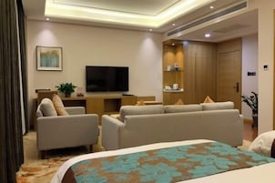 Quarto de hotel com cama, dois sofás, televisão e escrivaninha com planta.