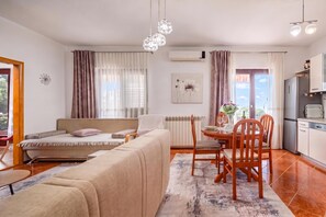 Uma sala de estar moderna com sofá, mesa de jantar com cadeiras, área de cozinha com geladeira e uma janela grande com cortinas.