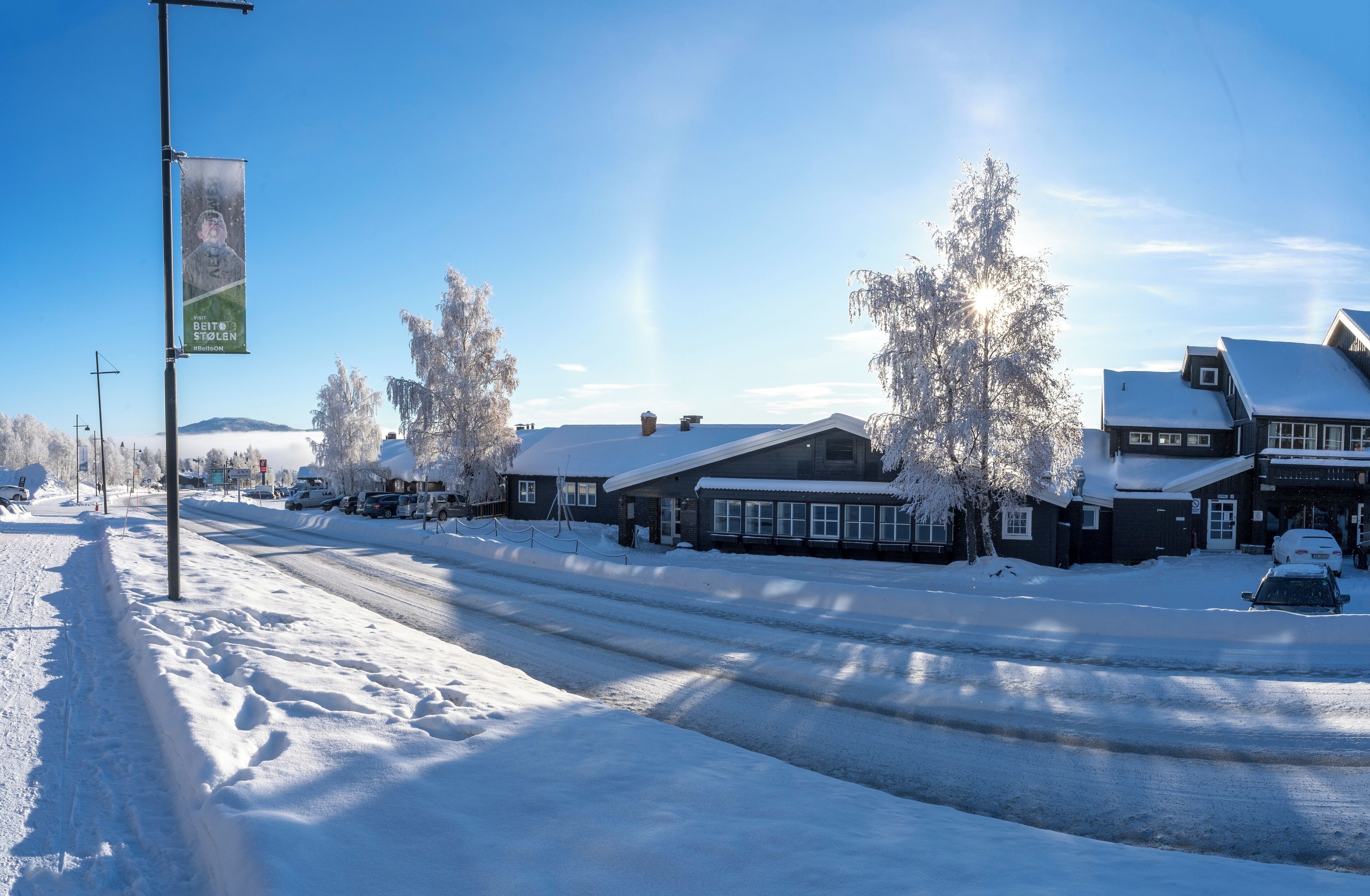 Rua coberta de neve com edifícios, incluindo uma casa com garagem e um prédio comercial com um letreiro.