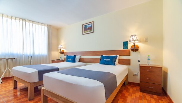 Quarto de hotel com duas camas, cabeceiras de madeira e mesa de cabeceira com abajur.