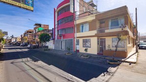 Um edifício de vários andares com fachada vermelha e bege, entrada protegida por grades e uma pequena árvore no jardim da frente.