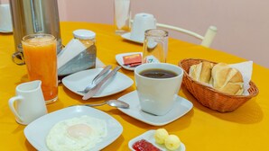 Mesa de café da manhã com xícara de café, copo de suco de laranja, prato com ovo, cesta de pão e pequeno ramequim de geleia.