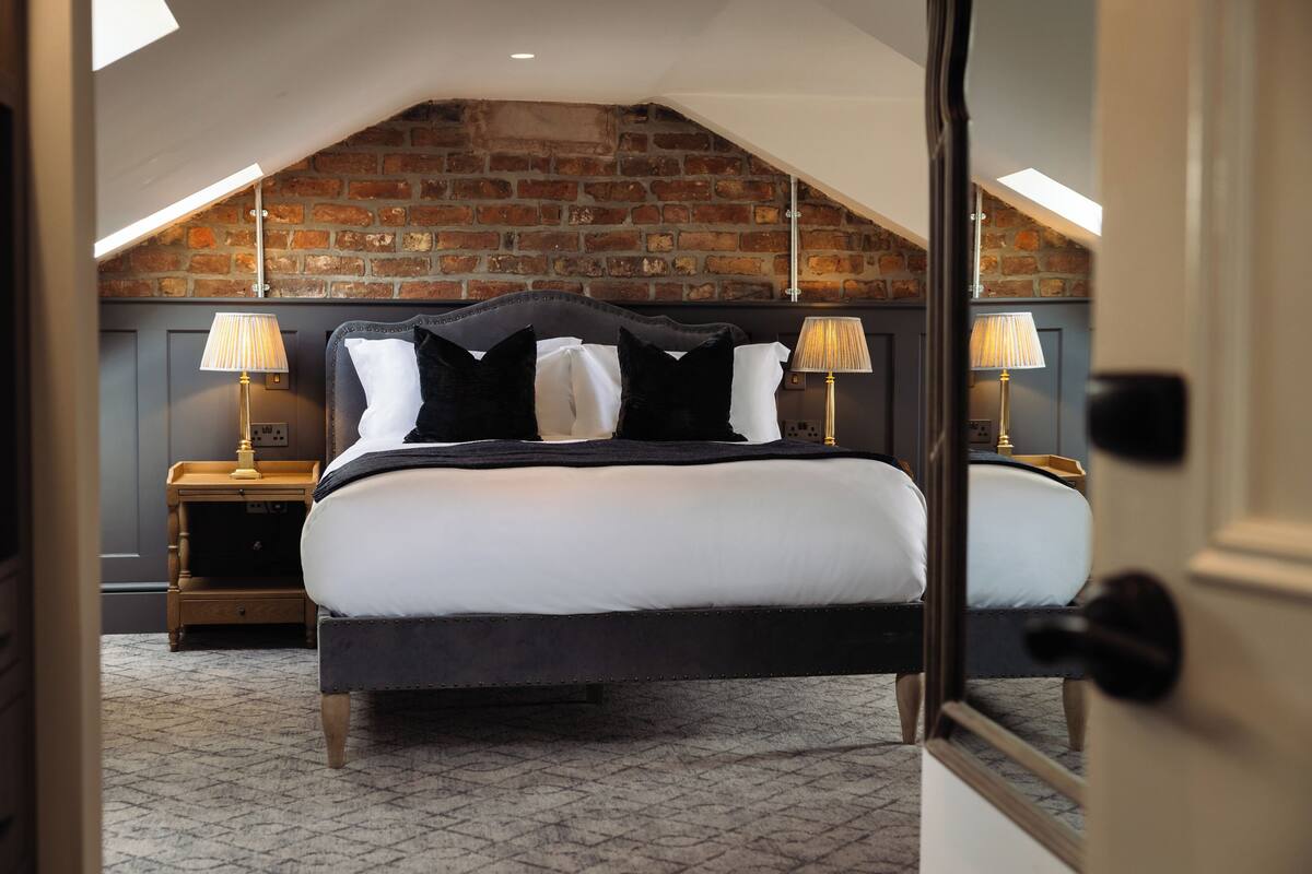 Quarto com parede de tijolos, teto inclinado, cama com roupa de cama preta e branca, duas mesas de cabeceira com luminárias e chão coberto por carpete.