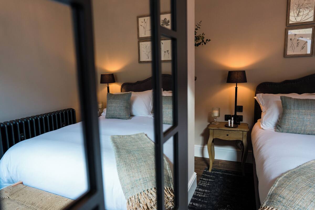 Quarto de hotel com duas camas, piso de madeira, radiador e arte botânica emoldurada na parede.
