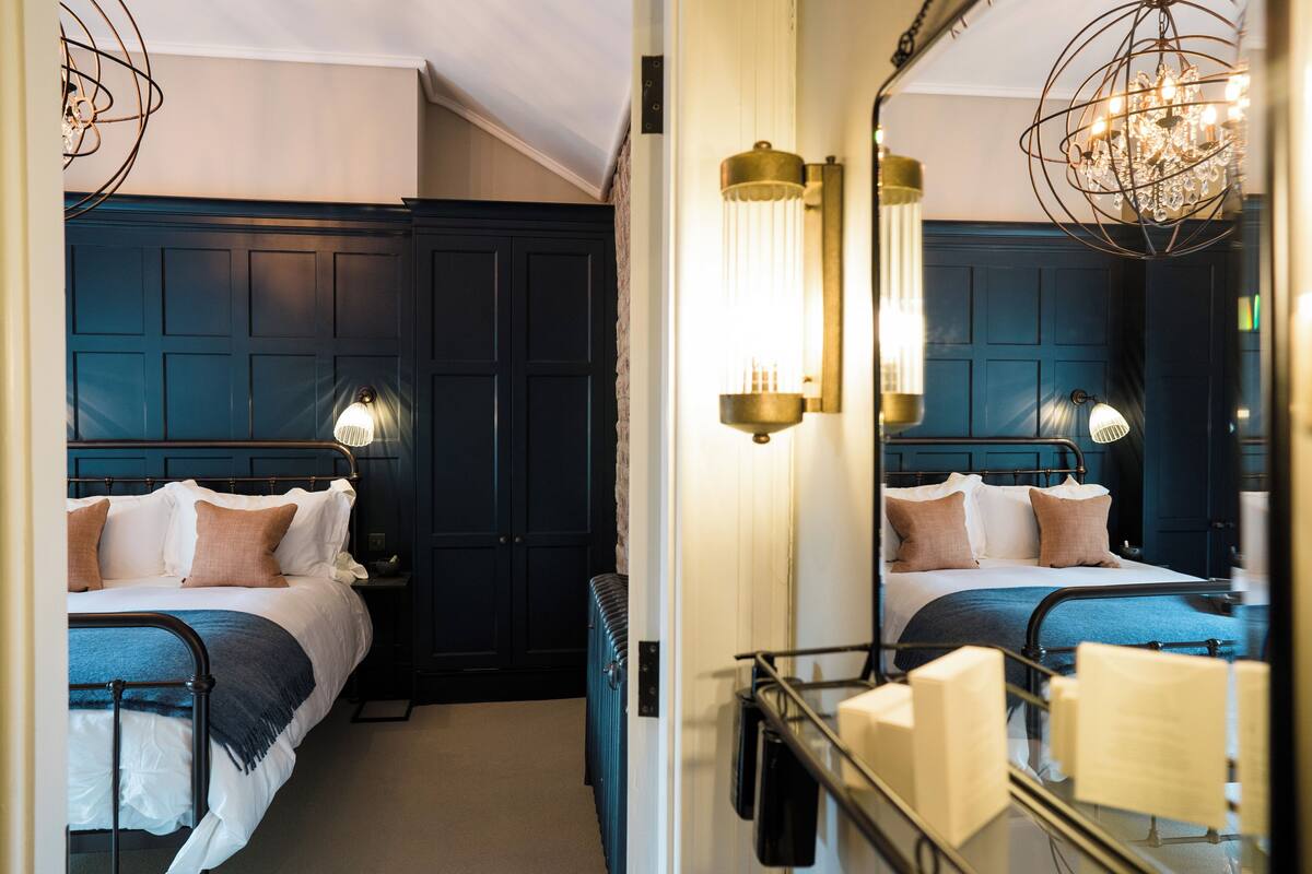 Quarto de hotel com parede azul escura, cama com roupa de cama branca, mesa de cabeceira com luminária e espelho com lustre.