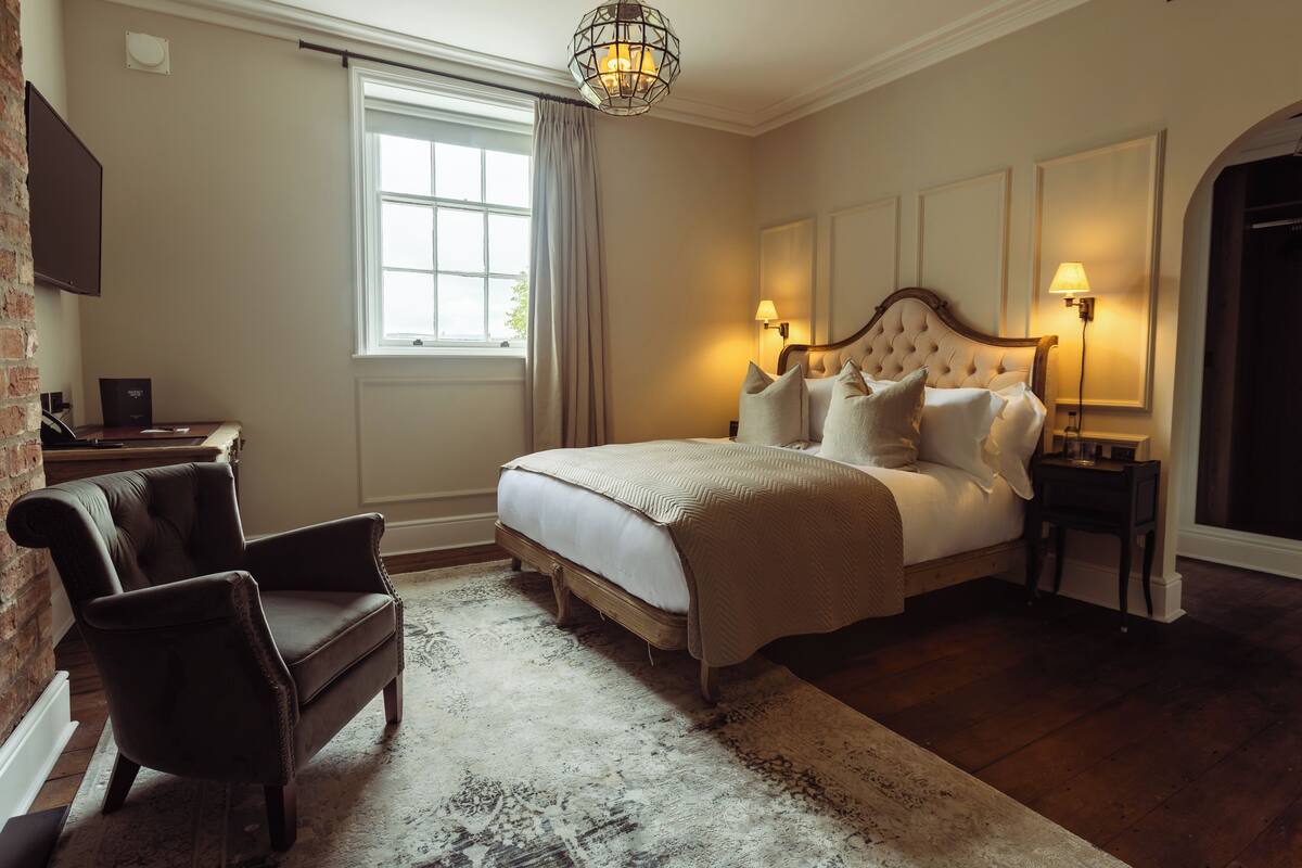 Quarto com cama grande, duas luminárias de cabeceira, uma cadeira, uma escrivaninha e uma janela com cortinas.