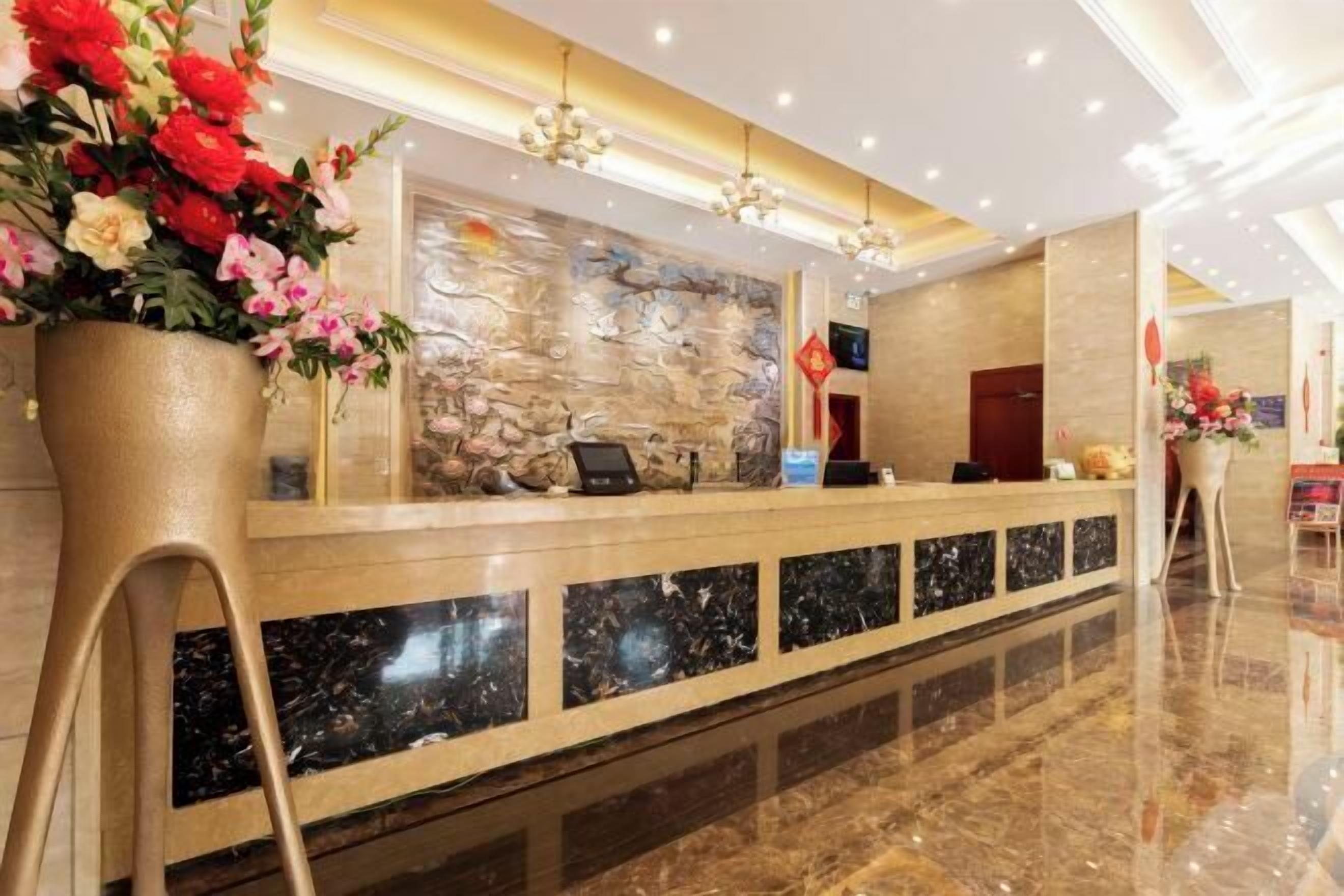 Uma área de recepção de hotel com balcão de mármore, parede de pedra com obras de arte e um vaso grande de flores.