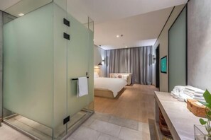 Quarto de hotel moderno com divisória de vidro, cama, banheiro com toucador e vista para a cidade.
