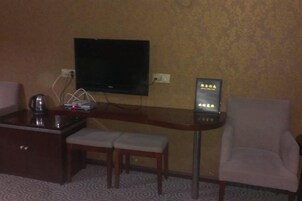 Quarto de hotel com televisão de tela plana na parede, uma pequena escrivaninha com um laptop, uma cadeira e um banco.