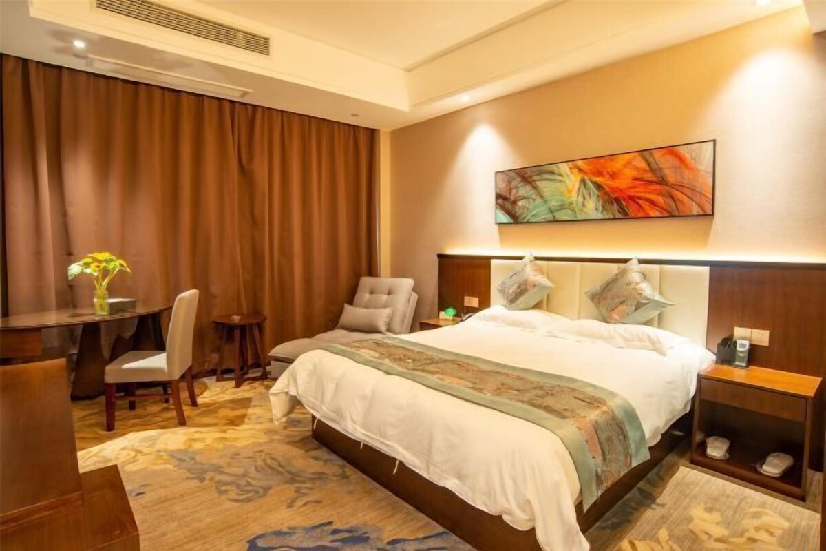 Quarto de hotel com uma cama grande, uma escrivaninha, uma cadeira, uma mesinha de cabeceira e uma obra de arte na parede.