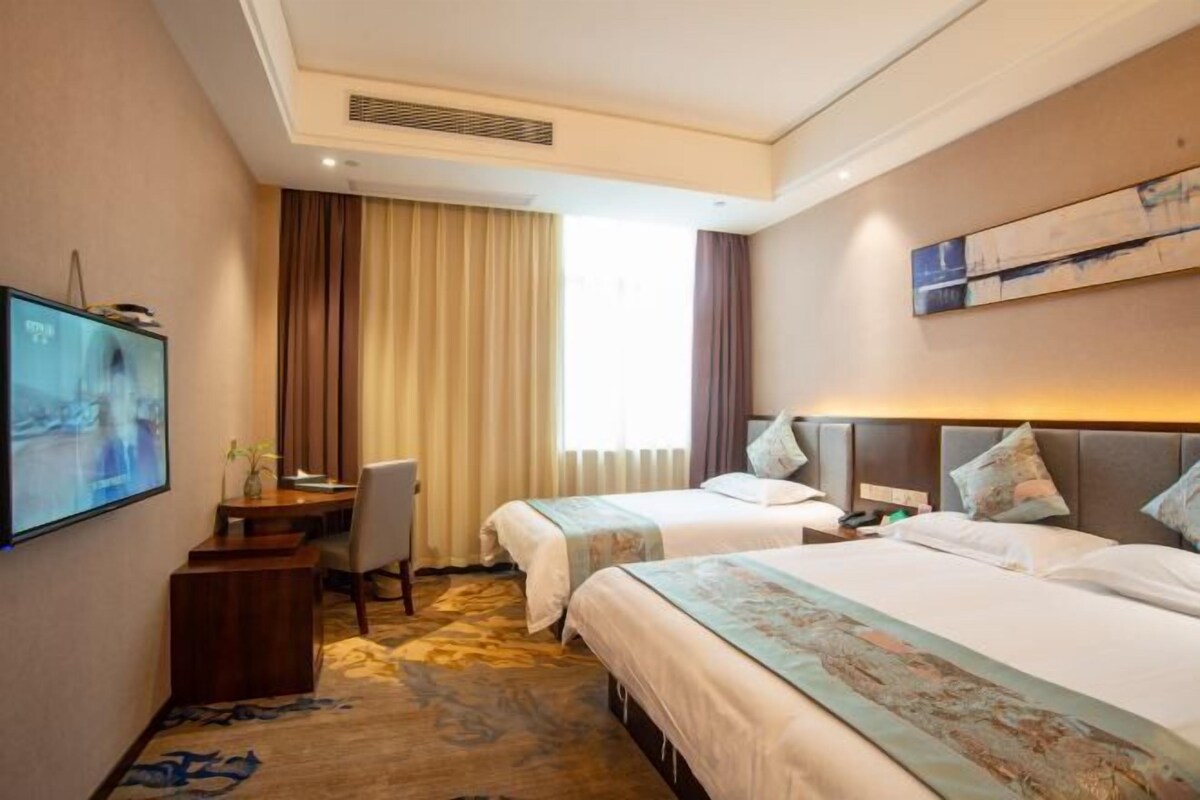 Quarto de hotel com duas camas, uma mesa, televisão e uma janela grande com cortinas.