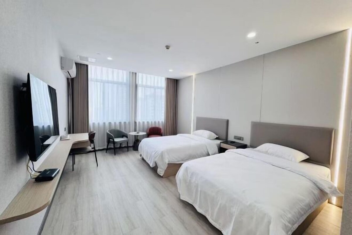 Quarto de hotel moderno com duas camas, uma escrivaninha, uma cadeira, televisão e janela com cortinas.
