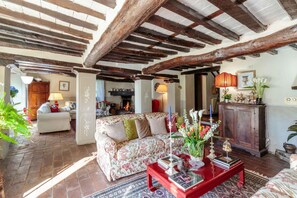 Uma sala de estar acolhedora com vigas de madeira aparentes, um sofá estampado com flores, uma mesa de centro vermelha e uma lareira ao fundo.