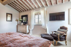 Quarto com cama, cômoda, cadeira, televisão e janela com cortinas.