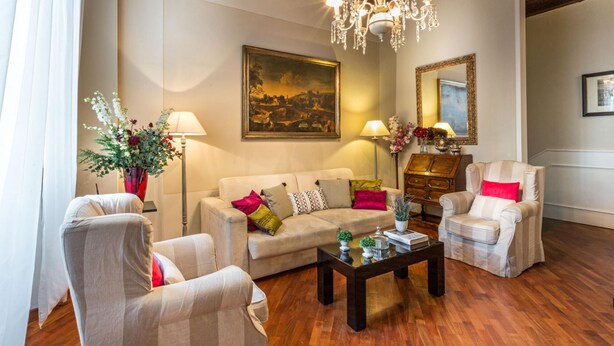 Uma sala de estar com um sofá bege, duas poltronas, uma mesa de centro, um candelabro, um quadro, um espelho e um vaso com flores.