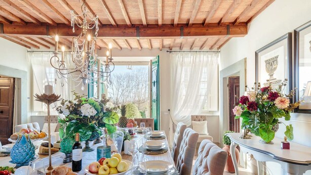 Uma sala de jantar com uma mesa de madeira posta para refeição, cercada por cadeiras, e decorada com flores e frutas.
