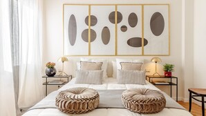 Quarto moderno com uma cama grande, duas mesas de cabeceira, uma obra de arte na parede e vista para o exterior.