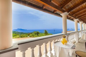 Varanda com vista para o mar e montanhas, balaústres brancos e uma mesa com um jarro amarelo.