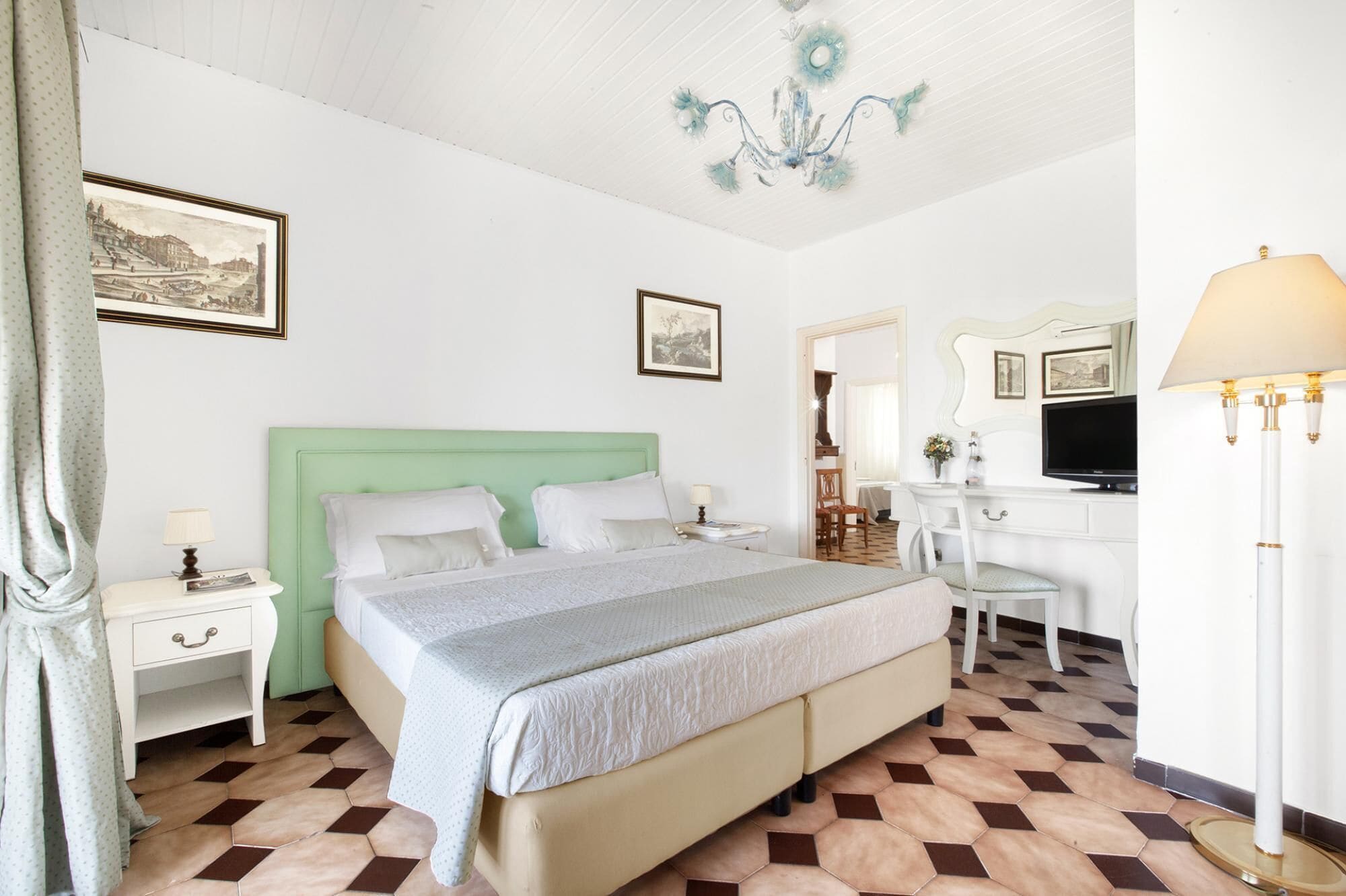 Quarto de hotel com cama, escrivaninha, televisão e lustre.