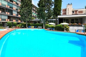 Piscina com espreguiçadeiras e guarda-sóis, cercada por vegetação e um edifício com varandas.