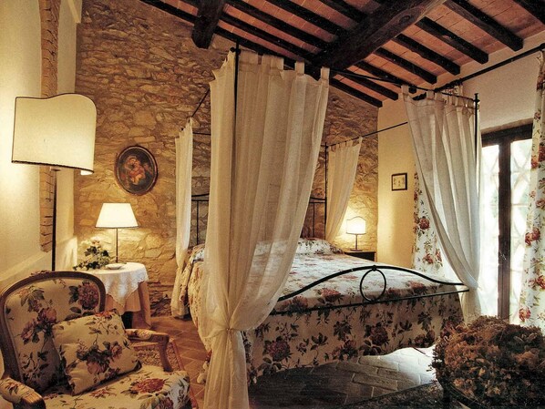 Quarto com parede de pedra, cama com dossel, poltrona floral e mesa de cabeceira com abajur.