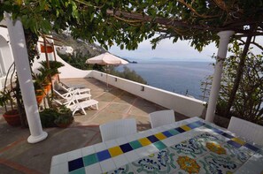 Um terraço com cadeiras brancas, uma mesa de azulejos e vista para o mar.