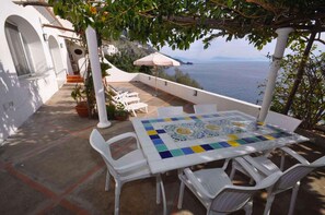 Um pátio externo com mesa de azulejos e cadeiras de plástico branco, com vista para um corpo d'água.