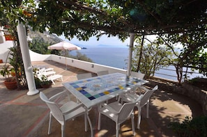 Um pátio com mesa de vidro e cadeiras de vime branco, com vista para um corpo d'água e uma paisagem montanhosa.