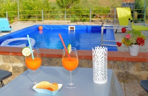 Cenário à beira da piscina com dois copos de bebida de laranja, um prato de petiscos e um vaso decorativo.