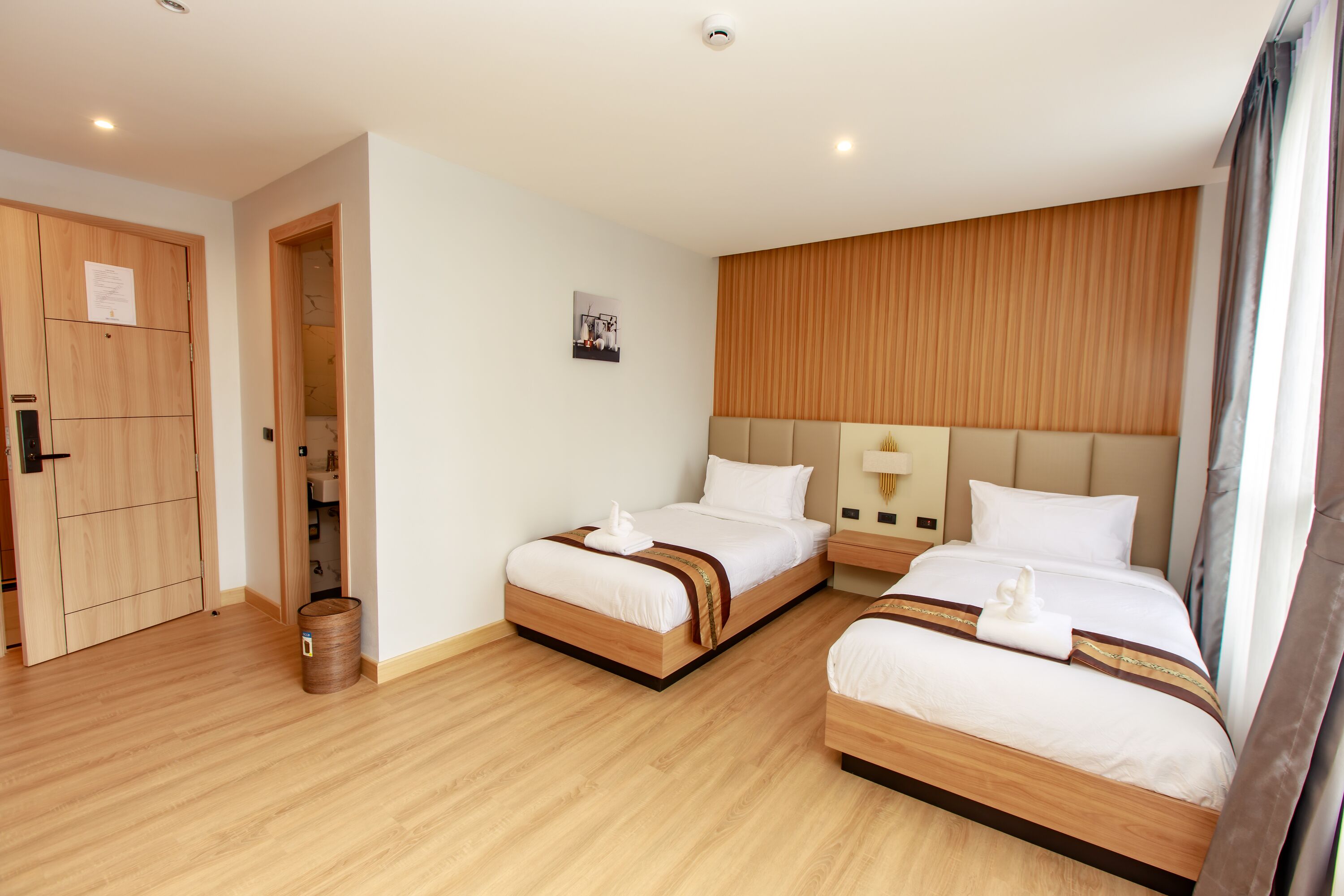 Quarto de hotel com duas camas, piso de madeira, painel de parede em madeira, mesa de cabeceira pequena com abajur e um quadro na parede.