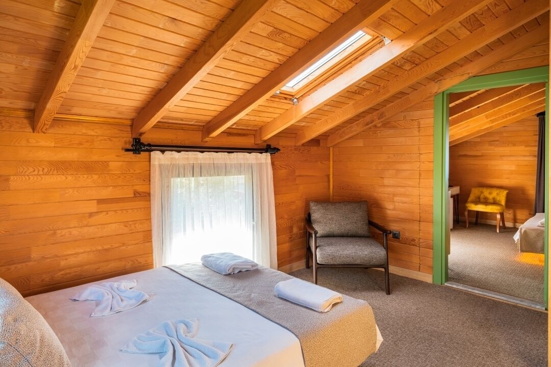 Quarto de cabana de madeira com uma cama, uma cadeira e vista através de uma porta aberta.