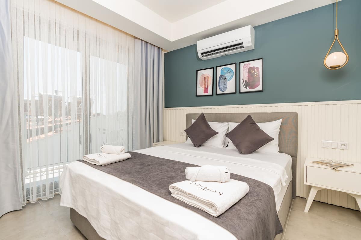 Quarto de hotel moderno com uma cama grande, roupa de cama branca, cabeceira cinza e vista da cidade através de cortinas leves.