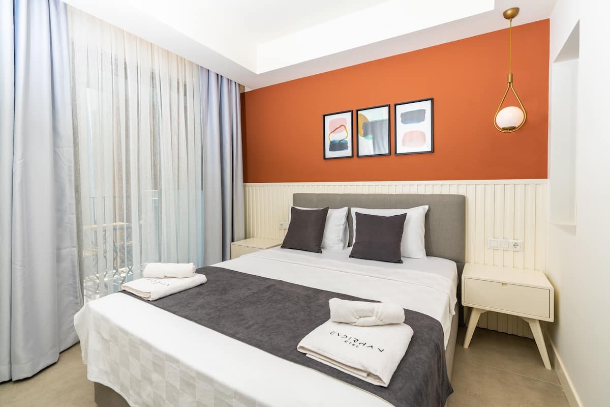 Quarto de hotel com cama, mesinhas de cabeceira brancas, janela com cortinas e uma parede com três quadros emoldurados.