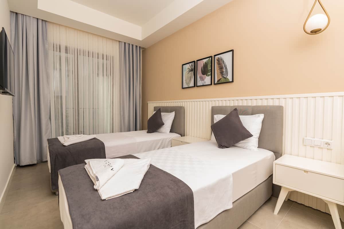 Quarto de hotel com duas camas, uma mesinha de cabeceira, televisão na parede e uma janela com cortinas.