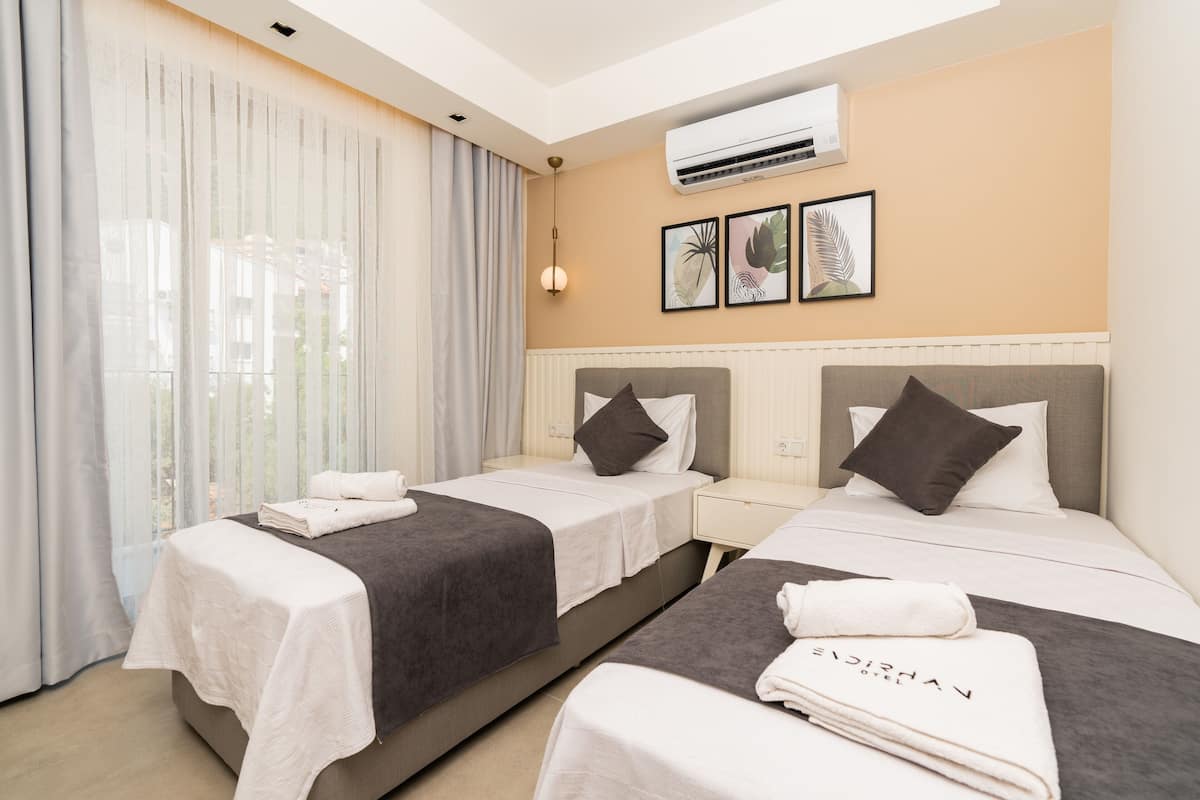 Quarto de hotel com duas camas, uma janela grande com cortinas leves, um ar condicionado e quadros decorativos na parede.