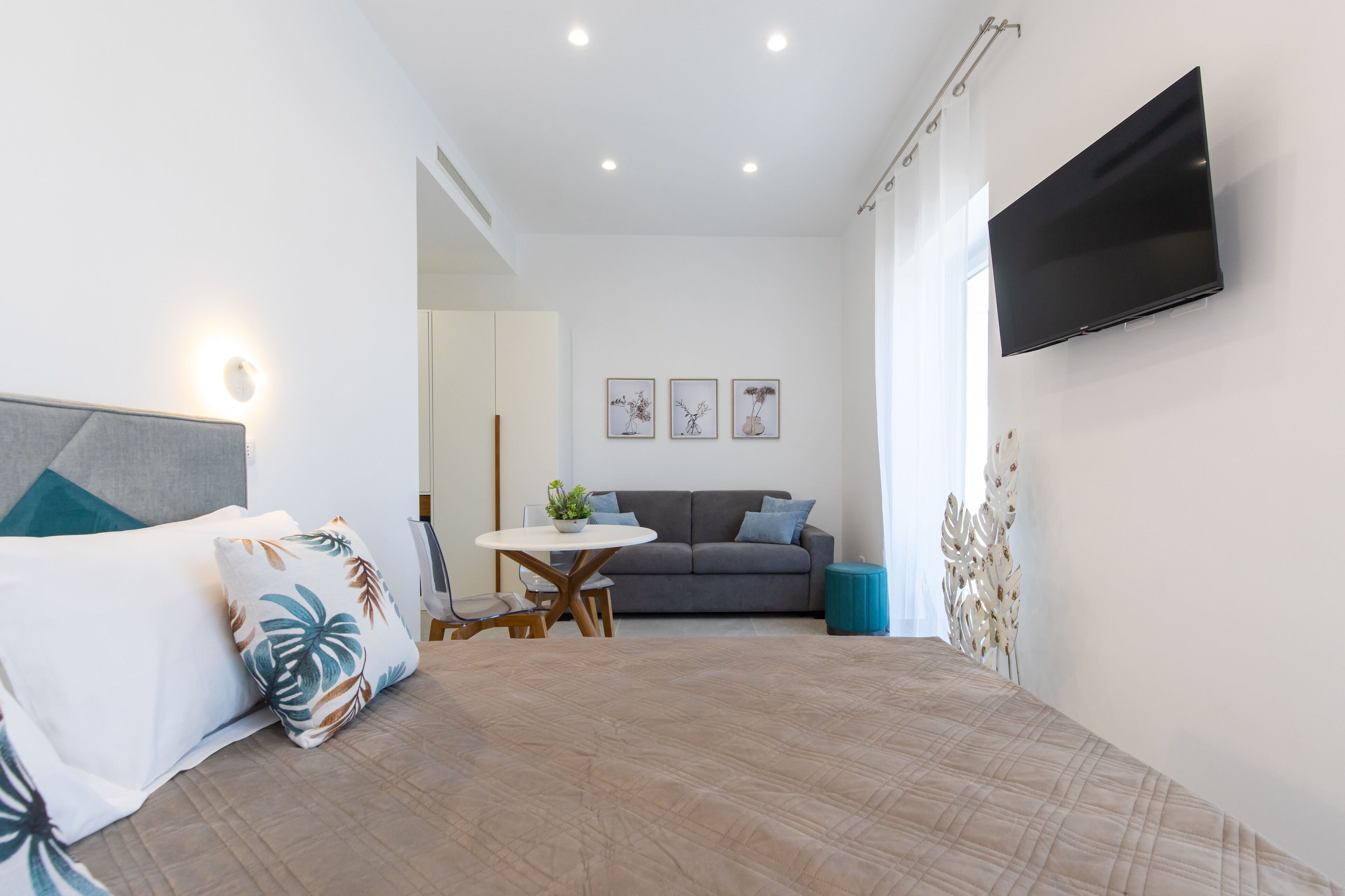 Quarto moderno com cama, televisão presa na parede, sofá e uma pequena mesa com uma planta.