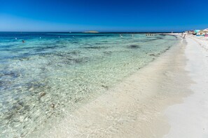 Uma praia com águas cristalinas e rasas, um areal e uma ilha ao longe.