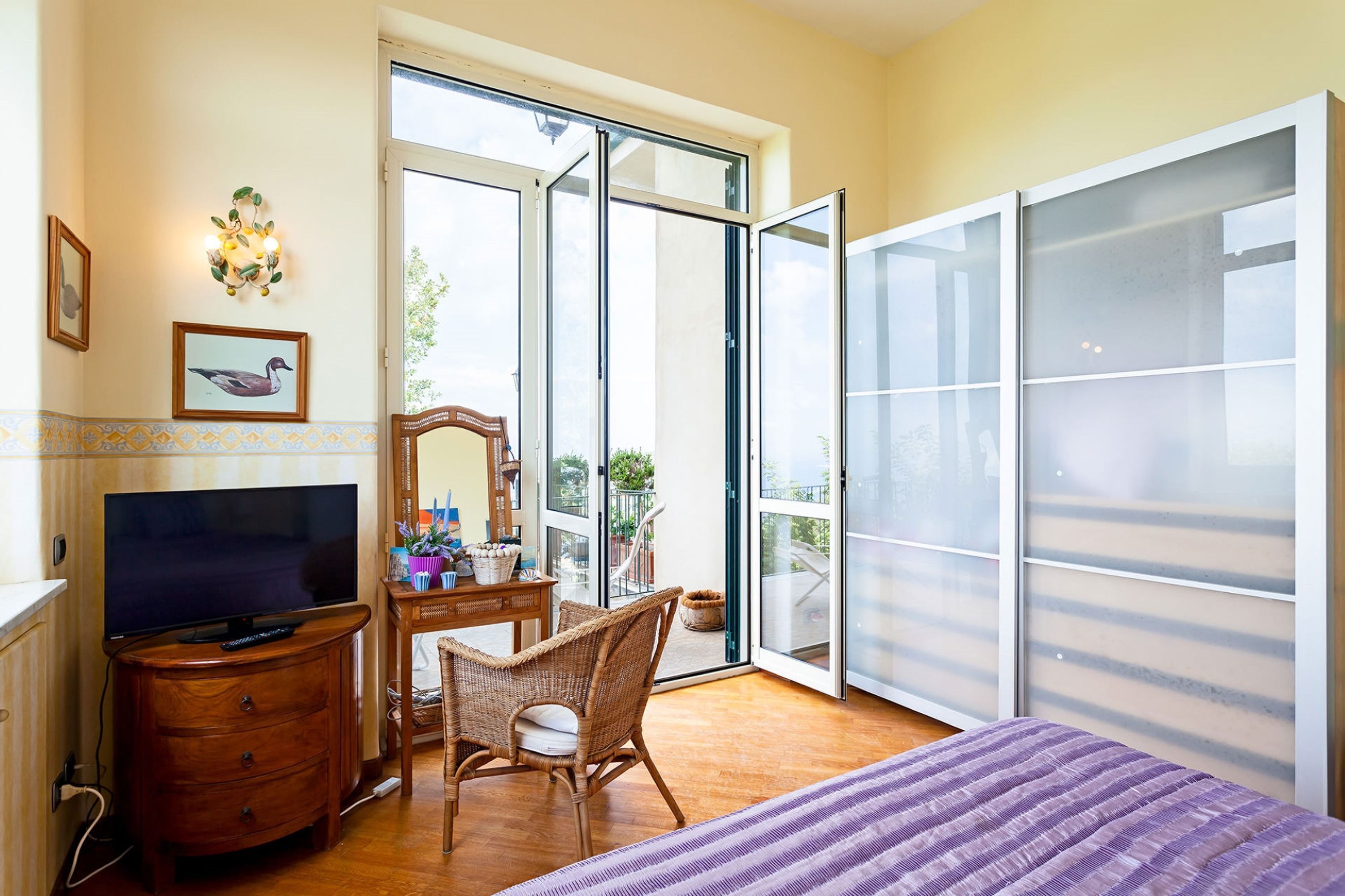 Quarto com cama, TV, escrivaninha com cadeira, penteadeira e porta de vidro deslizante que dá para uma varanda.