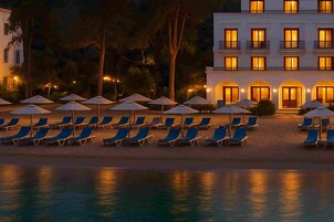 Um hotel à beira-mar com fileiras de espreguiçadeiras e guarda-sóis, uma piscina e um edifício com janelas iluminadas e varandas.