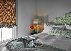 Cama bem arrumada com lençóis brancos, mesa de cabeceira com abajur, vaso com flores e janela com cortina estampada.