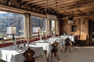 Uma sala de jantar com várias mesas preparadas para uma refeição, grandes janelas com vista para as montanhas e vigas de madeira no teto.